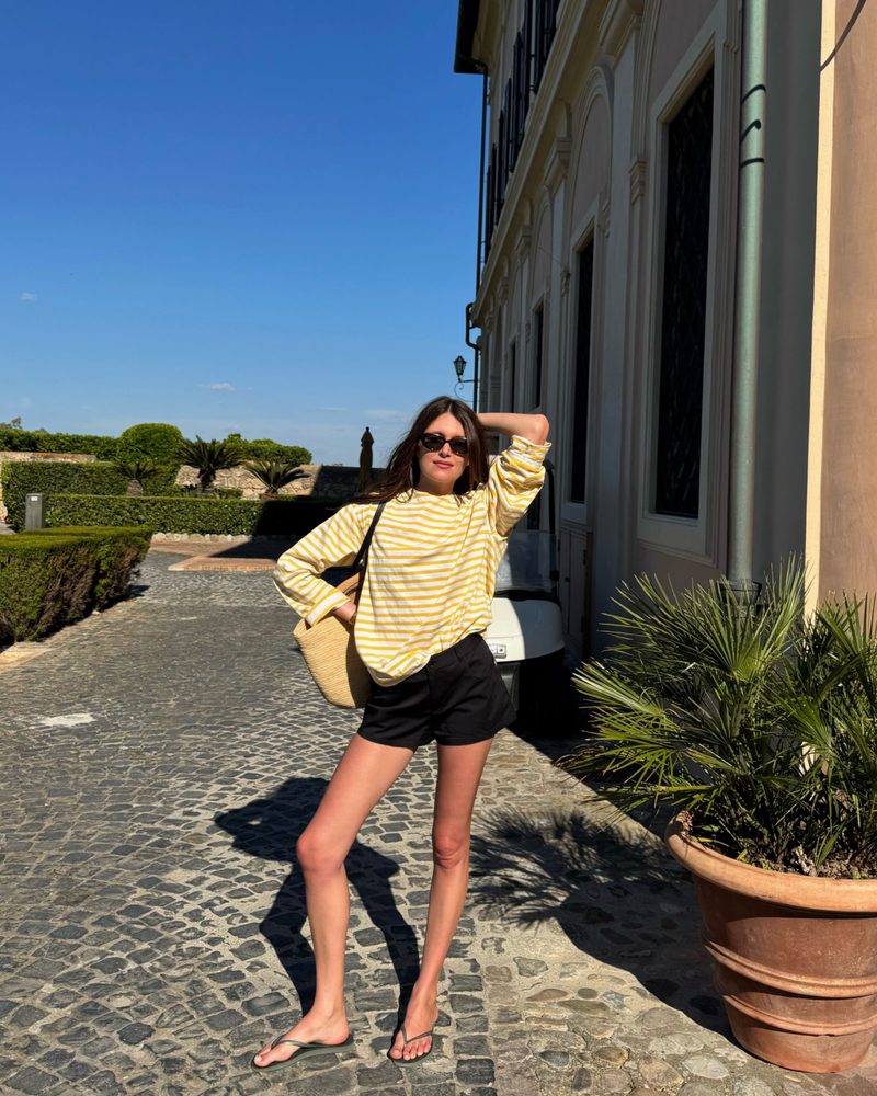 Yellow and white striped long-sleeve top paired with black shorts%2C accessorized with a woven tote bag and sunglasses%2C set against a sunny outdoor backdrop