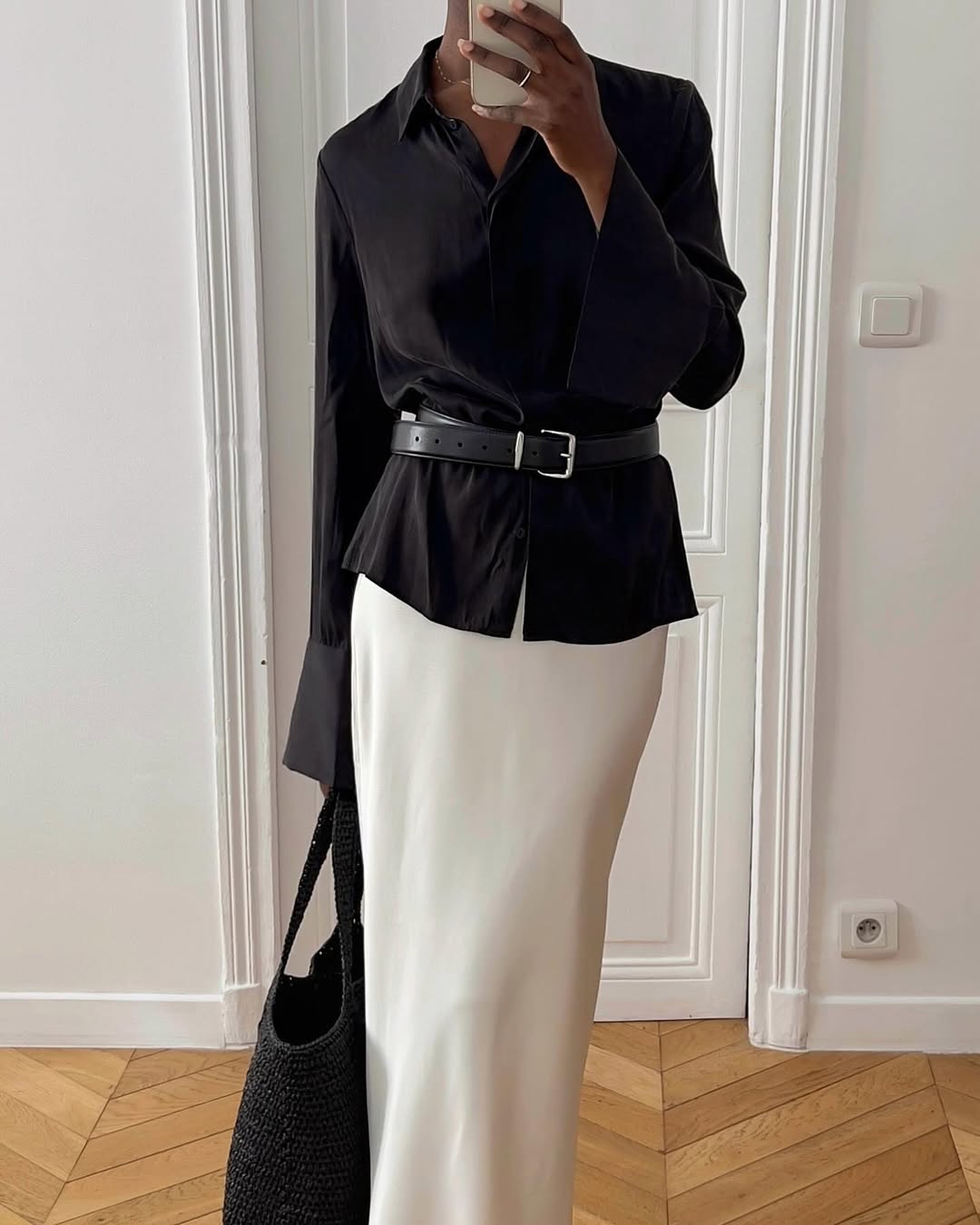 Chic outfit featuring a black silk shirt cinched with a wide leather belt%2C paired with a flowy white midi skirt and a textured black tote bag