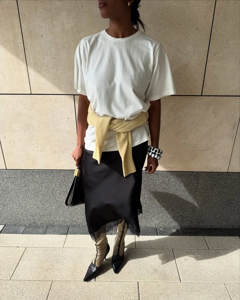 White crewneck T-shirt styled with a black satin slip skirt%2C knee-high boots%2C and a sweater tied at the waist for a minimal street style outfit