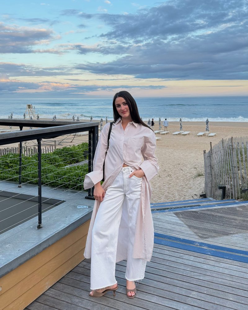 A woman poses on a wooden deck by the beach at sunset%2C wearing a striped shirt and white pants. The ocean and a cloudy sky form the backdrop.