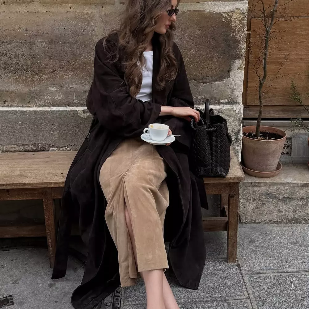 Chic autumn outfit featuring a brown suede long coat over a beige suede midi skirt, complemented by stylish black accessories and a coffee cup.