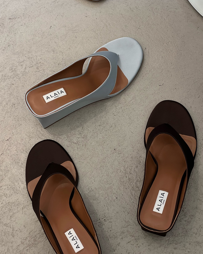 Ala%C3%AFa platform flip-flops in black and light gray%2C featuring a sleek cross-strap design and a chic wedge heel on a neutral backdrop.