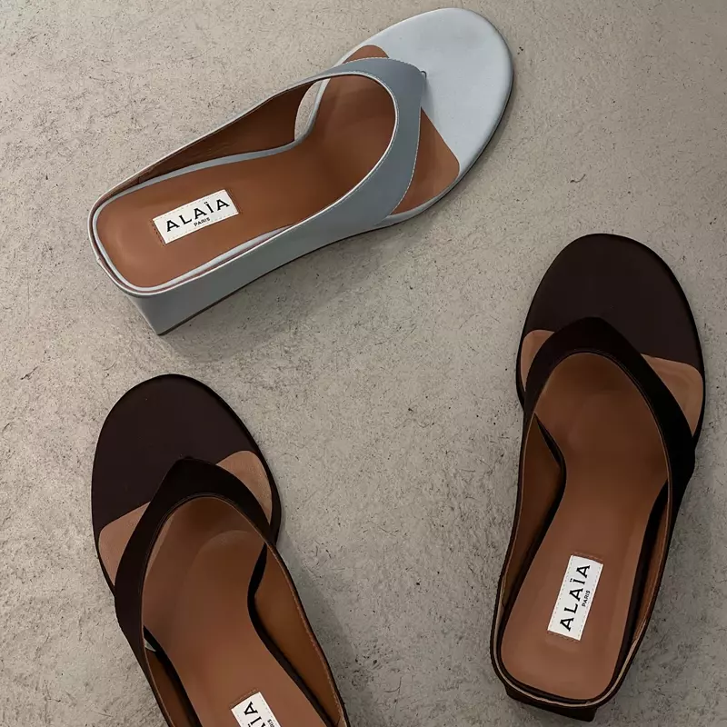 Alaïa platform flip-flops in black and light gray, featuring a sleek cross-strap design and a chic wedge heel on a neutral backdrop.