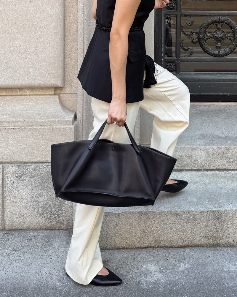 Oversized black leather work tote carried with a sleeveless black top%2C cream trousers and pointed flats on stone steps