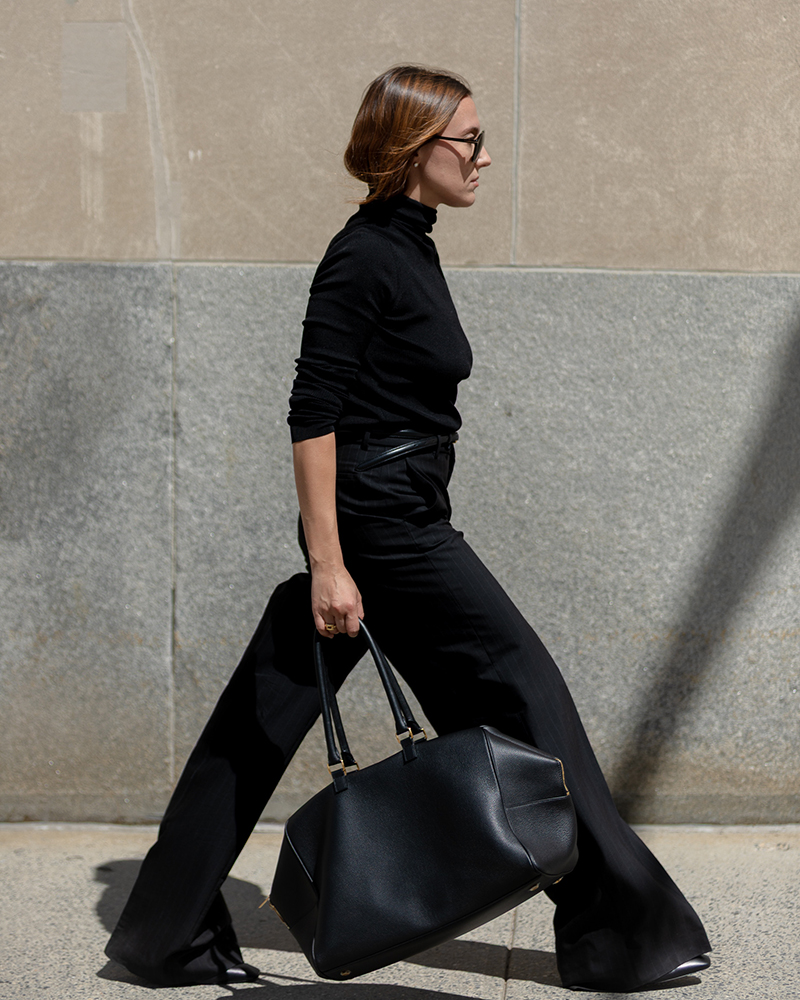 Street-style look with black turtleneck and wide-leg trousers%2C carrying a structured black leather tote past a stone wall
