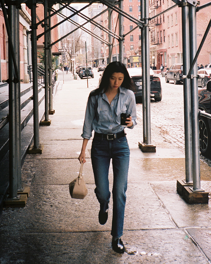 Light blue button-down shirt styled with straight-leg jeans%2C black loafers and a beige handbag on a city sidewalk