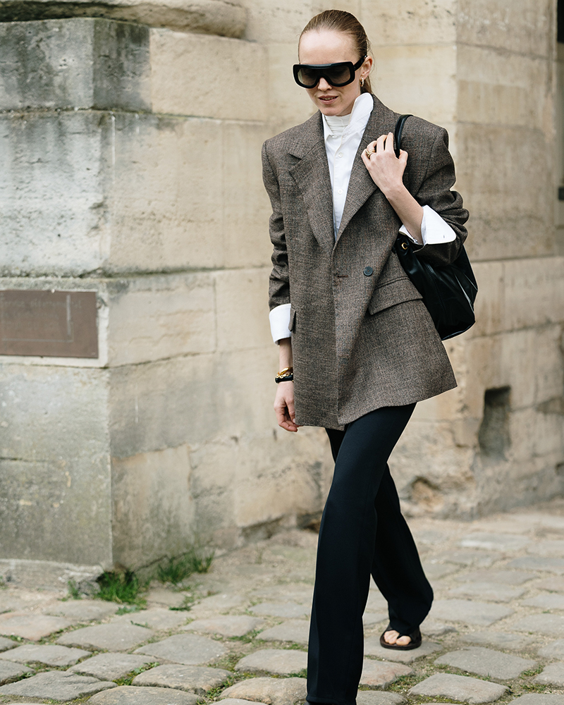Model in an oversized brown tweed blazer%2C white shirt and black trousers with a leather shoulder bag on cobblestones