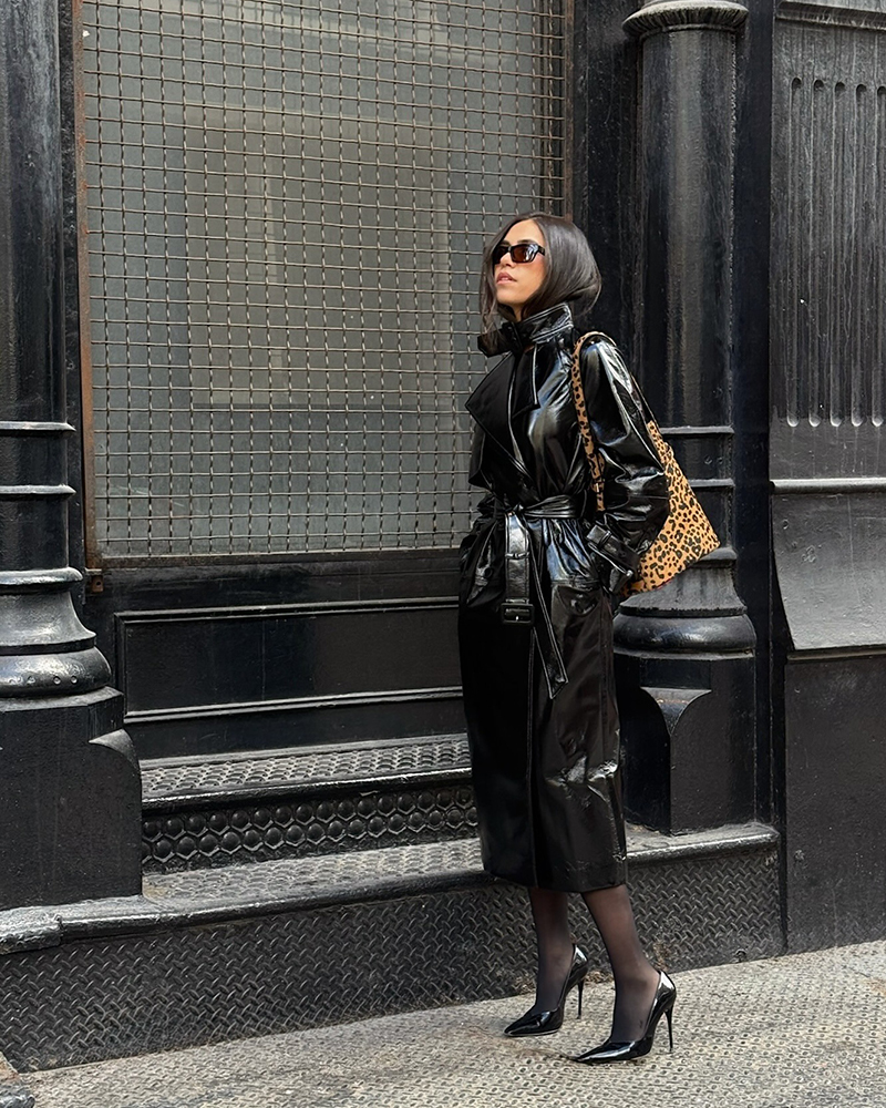 Street style outfit in glossy black belted trench coat%2C sheer tights and patent heels with leopard shoulder bag on dark city steps
