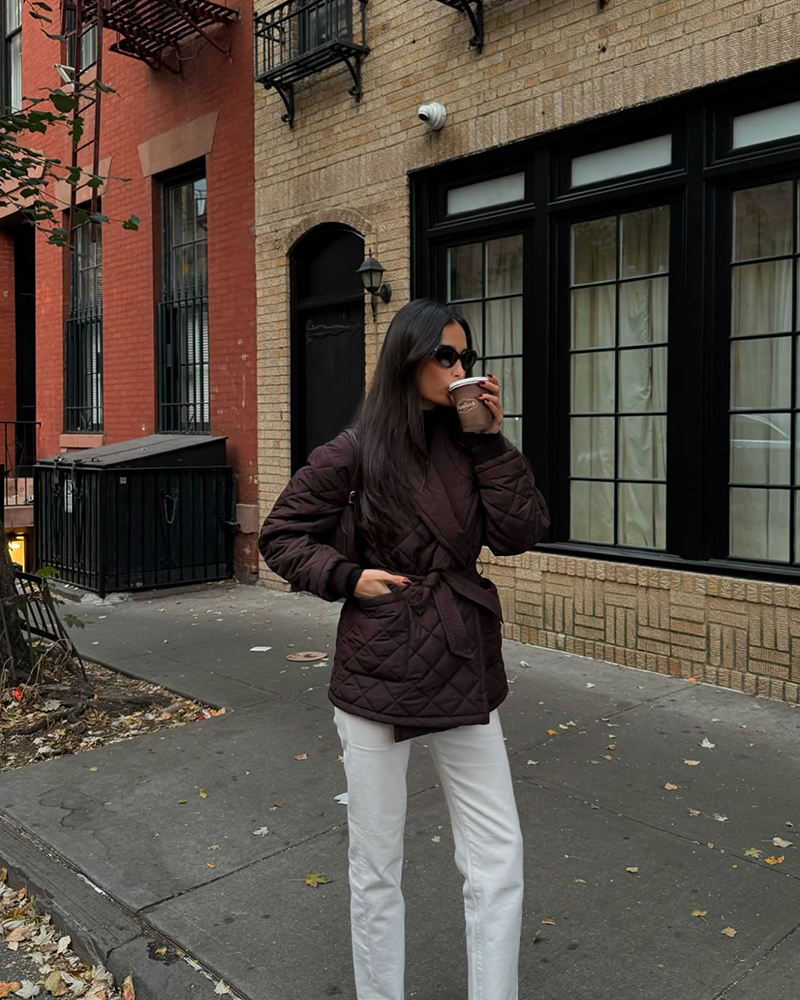 Street style look featuring a chocolate brown quilted jacket with tie waist%2C white straight-leg jeans and sunglasses%2C styled on a city sidewalk with brick buildings