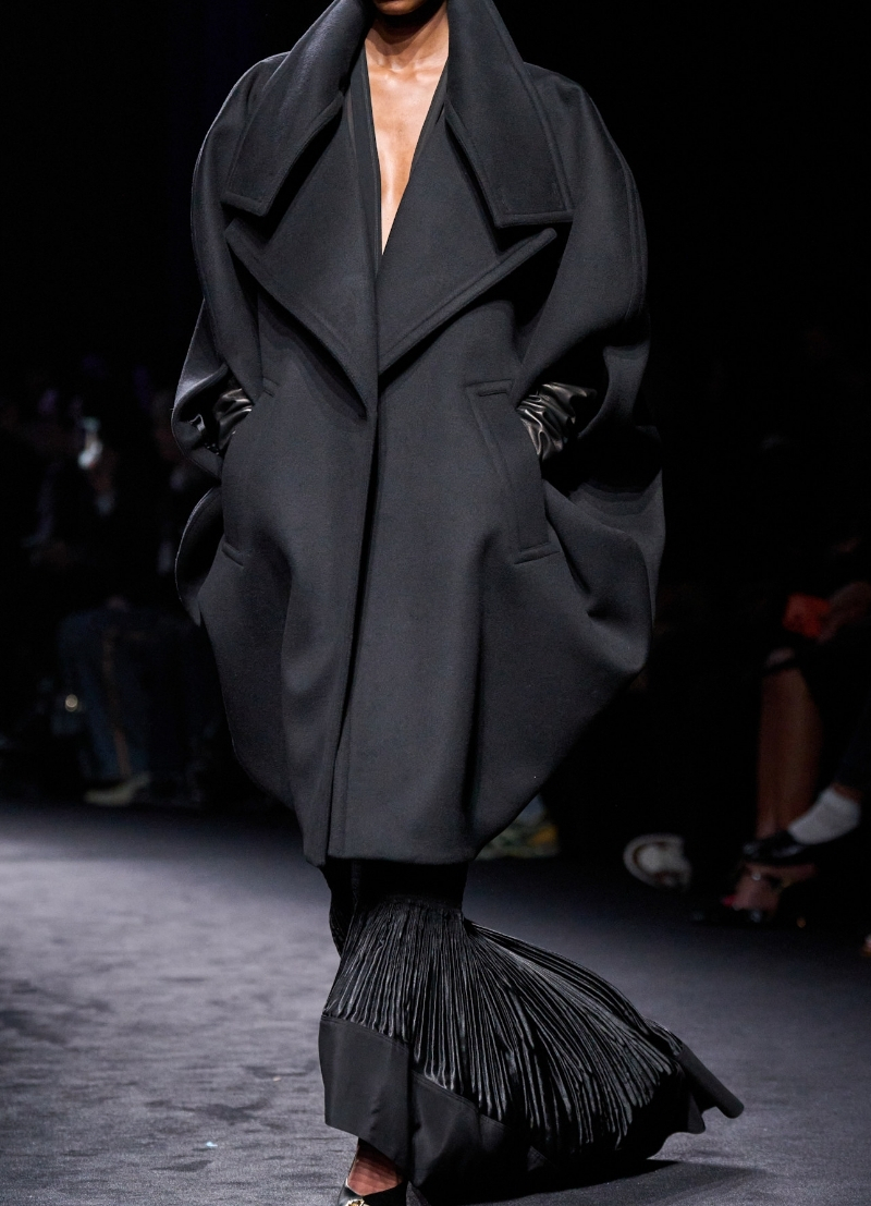 Oversized black wool coat with dramatic lapels and sculptural silhouette styled with a pleated skirt on the runway