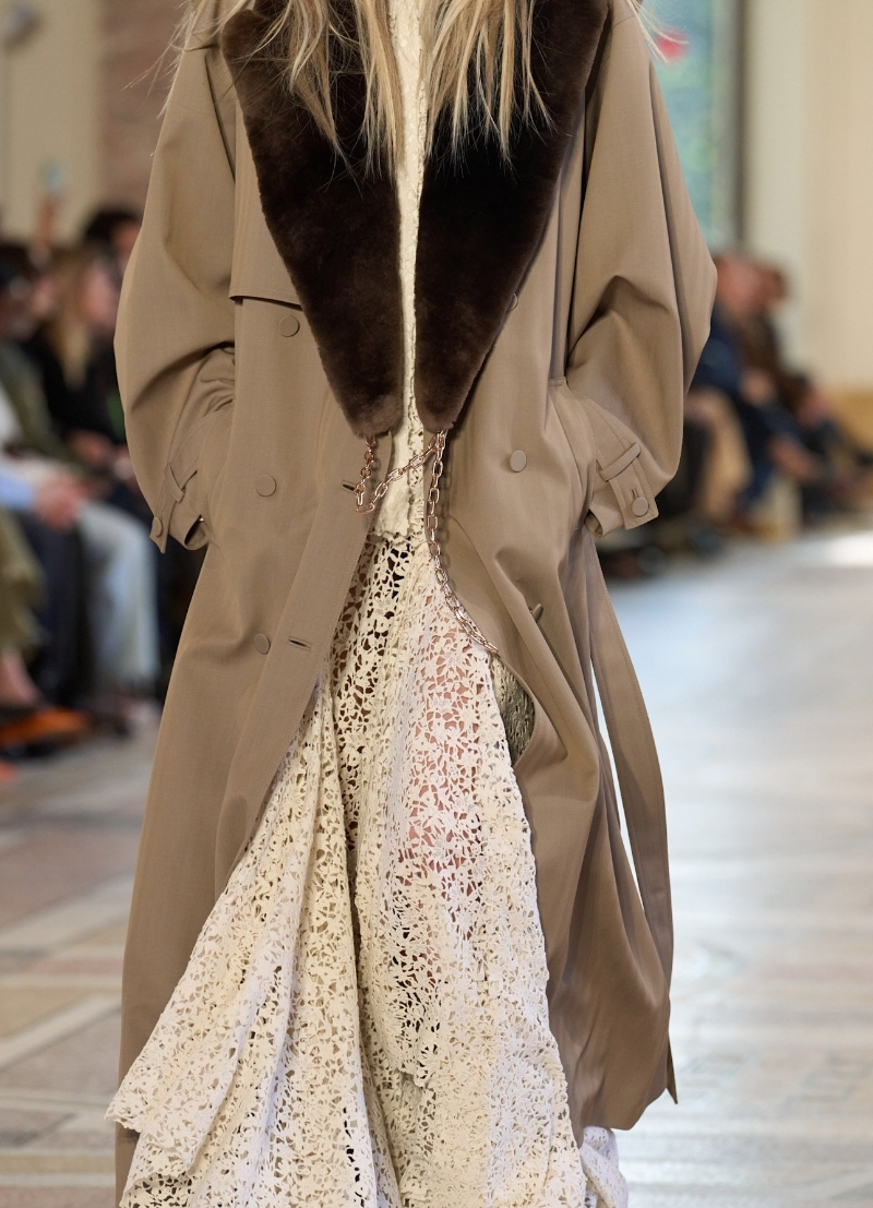 Taupe oversized trench coat with dark brown fur collar layered over a cream lace dress on the runway
