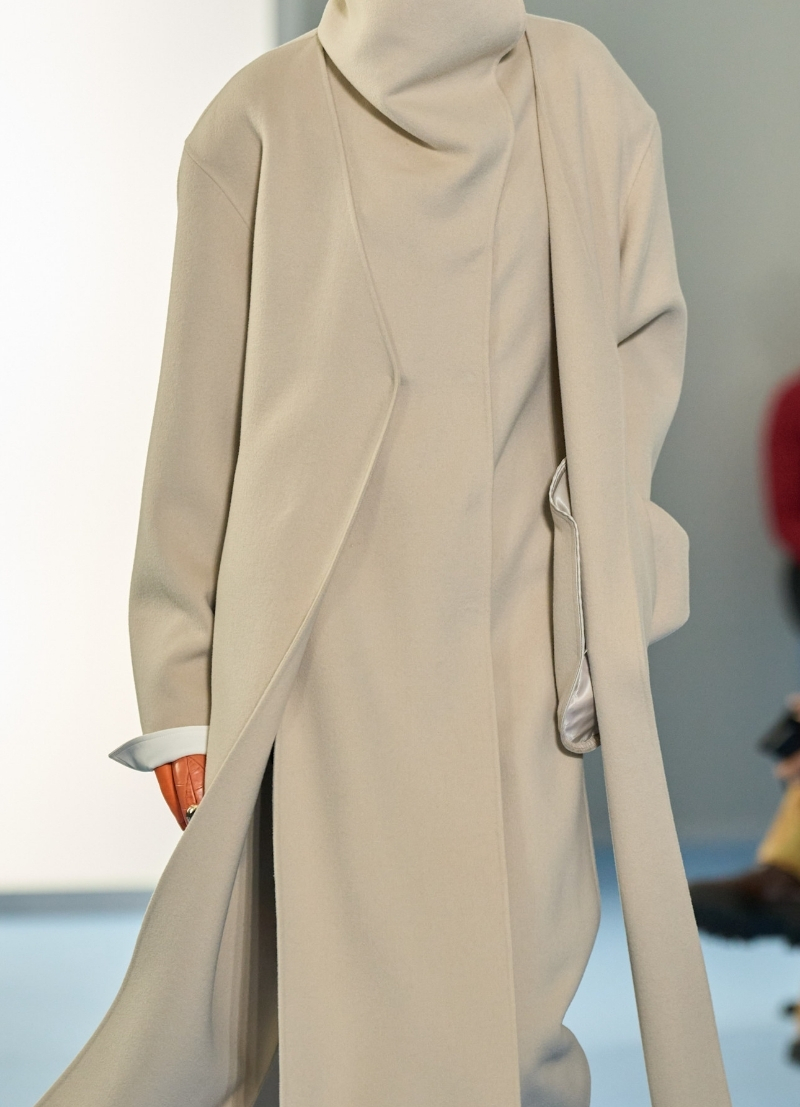 Minimalist cream long coat with draped high neck and elongated scarf detail presented on the runway