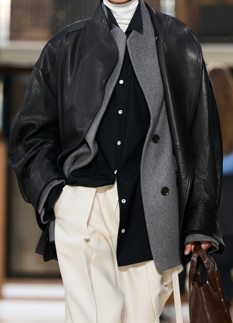 Oversized black leather jacket layered with a gray coat, black shirt and cream trousers on the runway