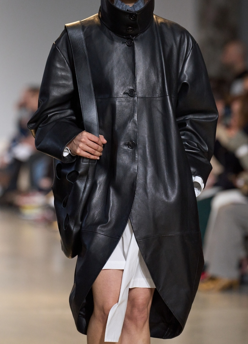 Black leather cocoon coat with high collar and shoulder bag styled over a white dress on the runway