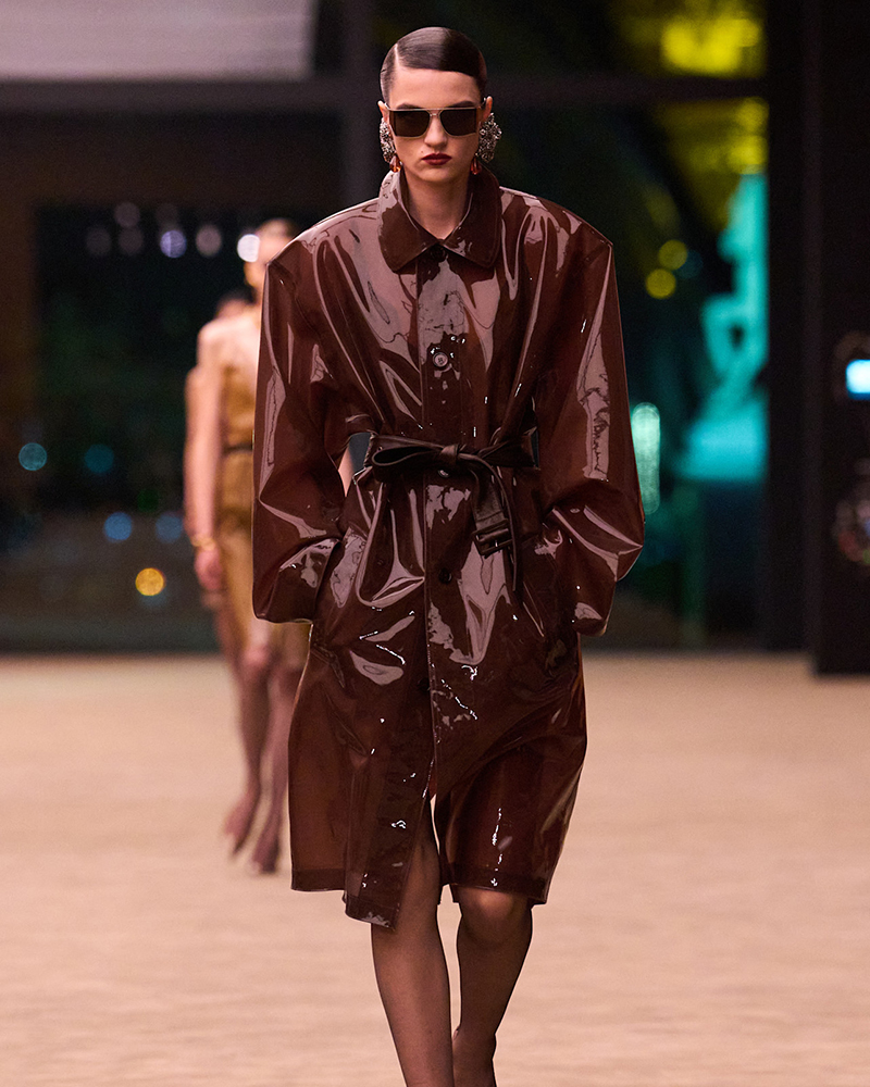 Model in a glossy chocolate brown belted trench coat with sunglasses and statement earrings on the runway