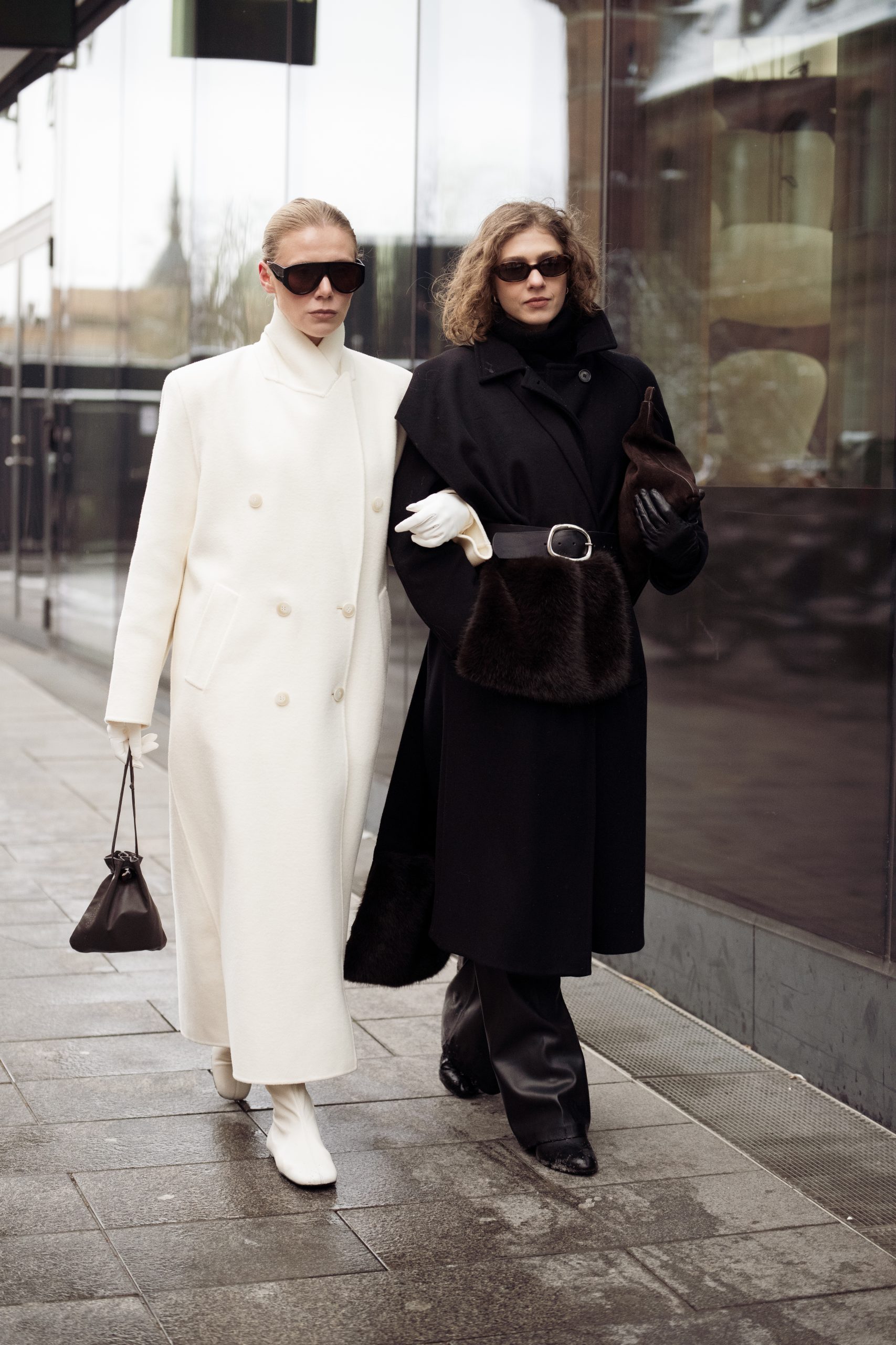 Chic street style at Copenhagen Fashion Week features a model in a white double-breasted coat and another in a black belted cape%2C both paired with oversized sunglasses and sleek boots.