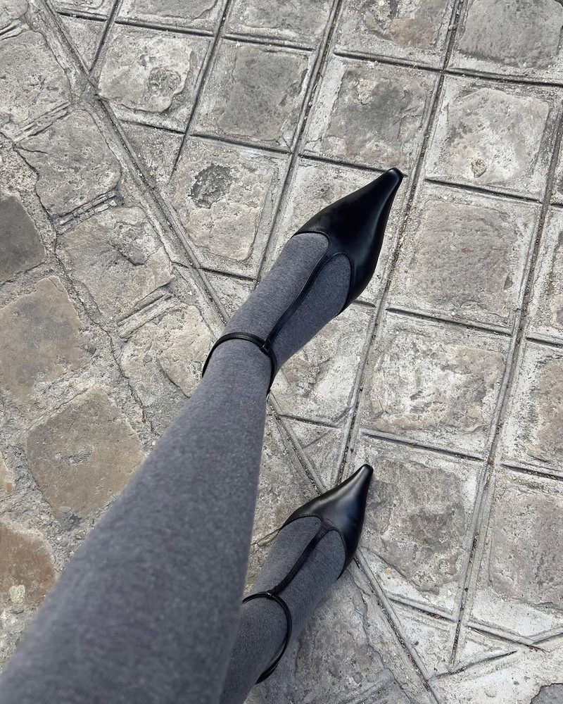 Black leather pointed toe shoes with elastic slingback straps styled over gray tights on textured stone pavement