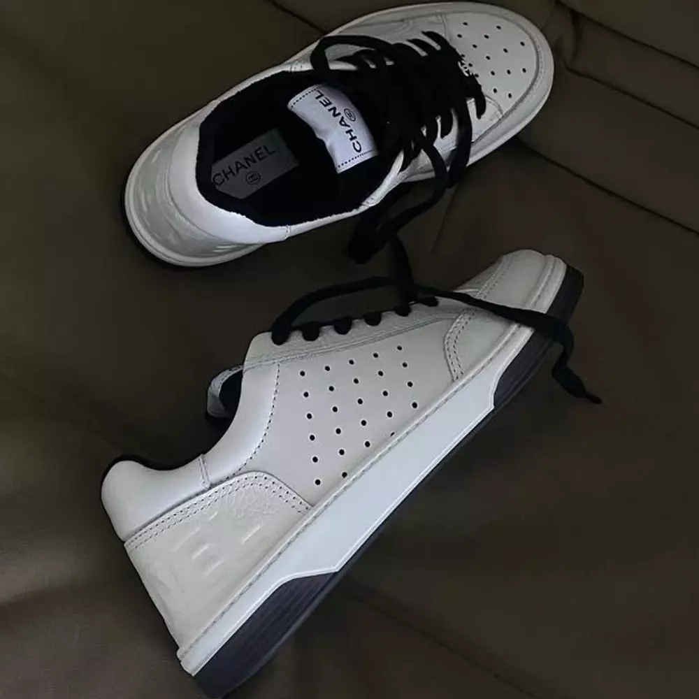 Chanel white sneakers with black laces feature a sleek design and perforated detailing, elegantly placed on a soft beige chair