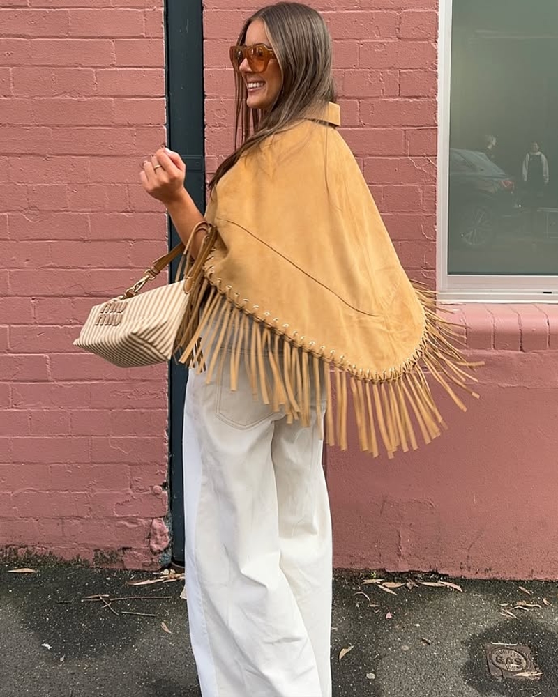 Fringe suede poncho in a warm tan hue%2C styled with high-waisted white trousers and a chic woven bag%2C perfect for a trendy fall look.