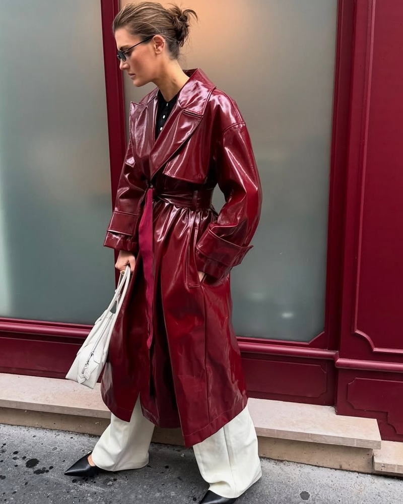 Bold burgundy patent leather trench coat cinched at the waist%2C paired with cream wide-leg trousers and a chic white handbag%2C embodying fall elegance