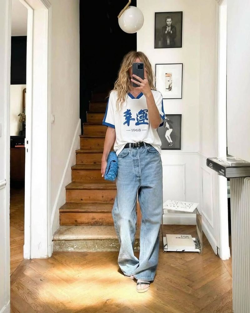 A person in a white and blue shirt with jeans takes a mirror selfie in a stylish hallway. They hold a blue clutch and stand by a wooden staircase.