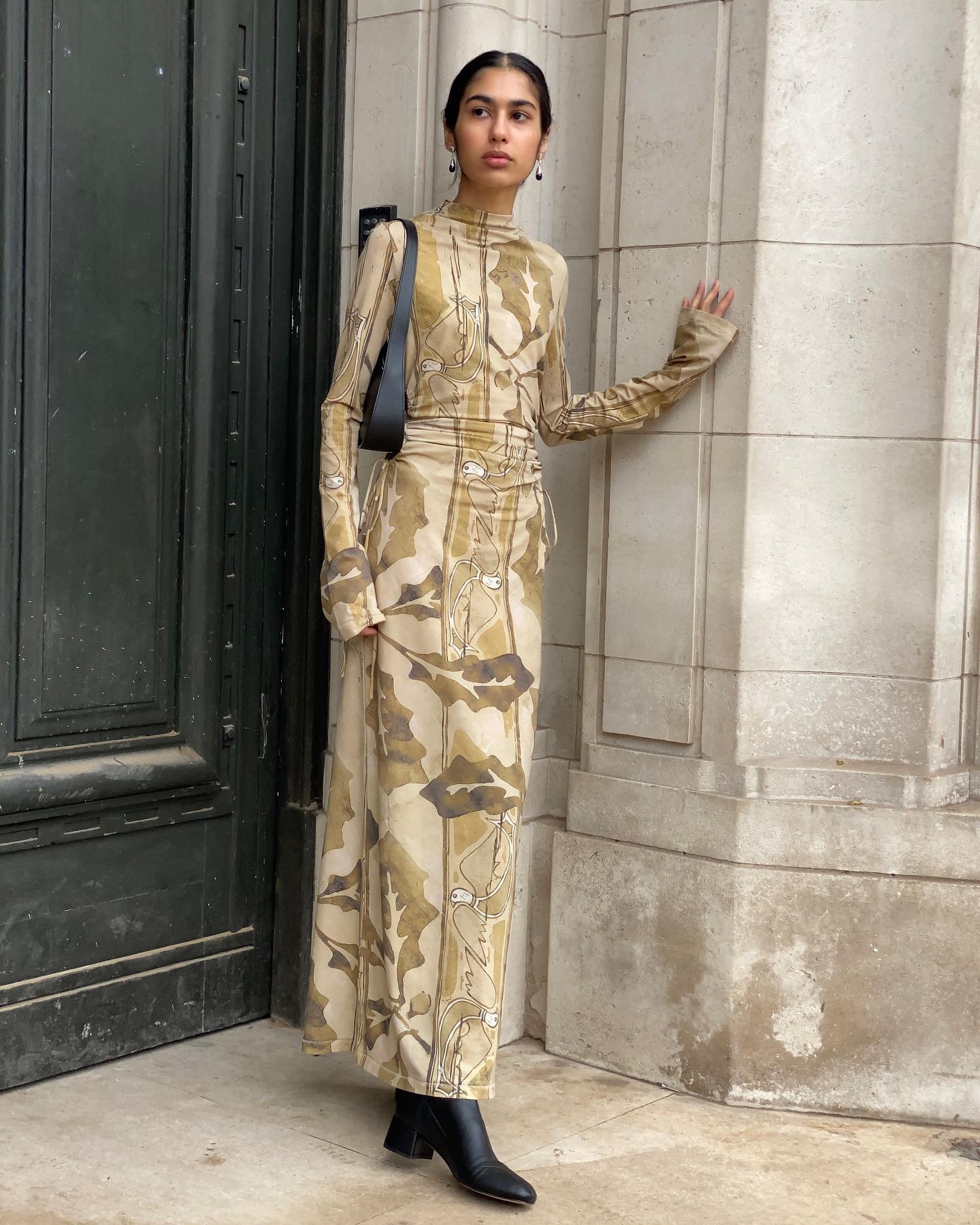 Model wearing a long-sleeve beige and olive abstract print maxi dress paired with black ankle boots and a black shoulder bag