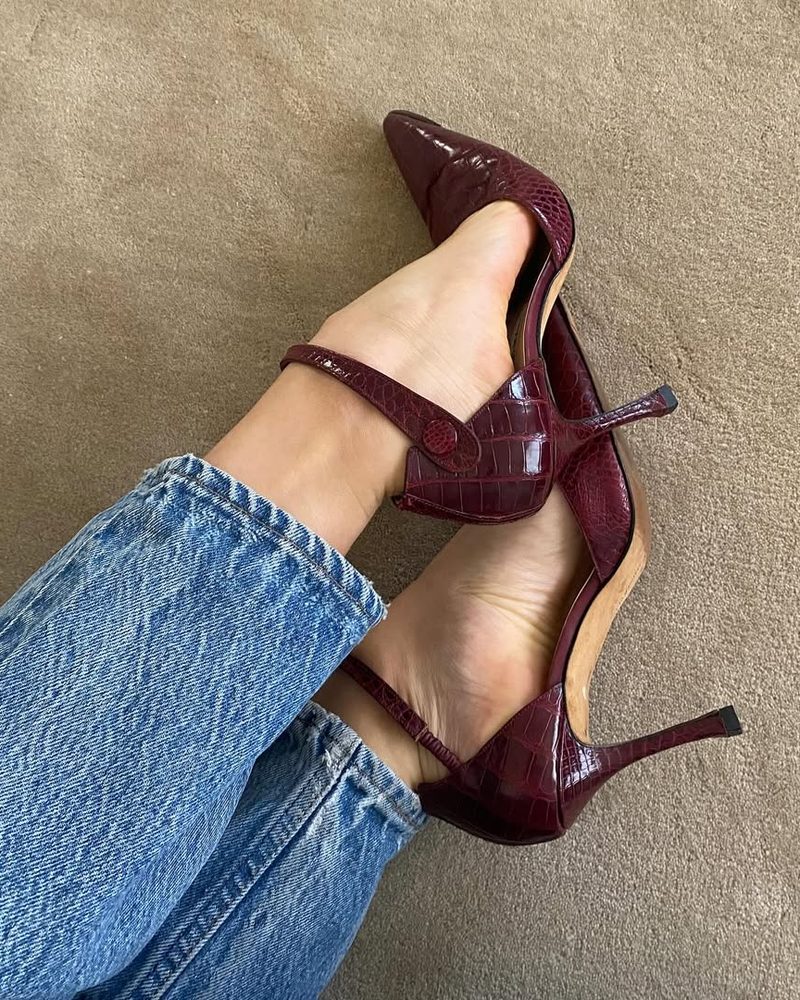 Woman wearing stylish burgundy crocodile-embossed high heel pumps with ankle straps%2C paired with blue denim jeans%2C resting on a beige carpet.