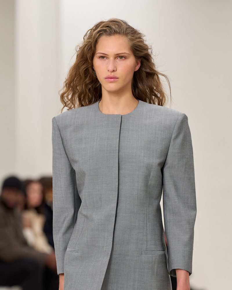 A model with wavy hair walks the runway wearing a minimalist gray structured blazer with sharp shoulders%2C set in a sleek fashion show environment.