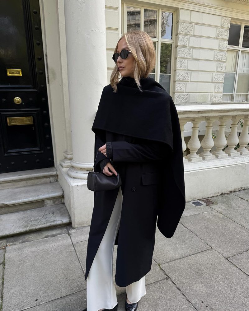 Sleek black cape coat layered over a fitted black top%2C paired with wide-leg cream trousers and glossy black loafers%2C exuding modern elegance.