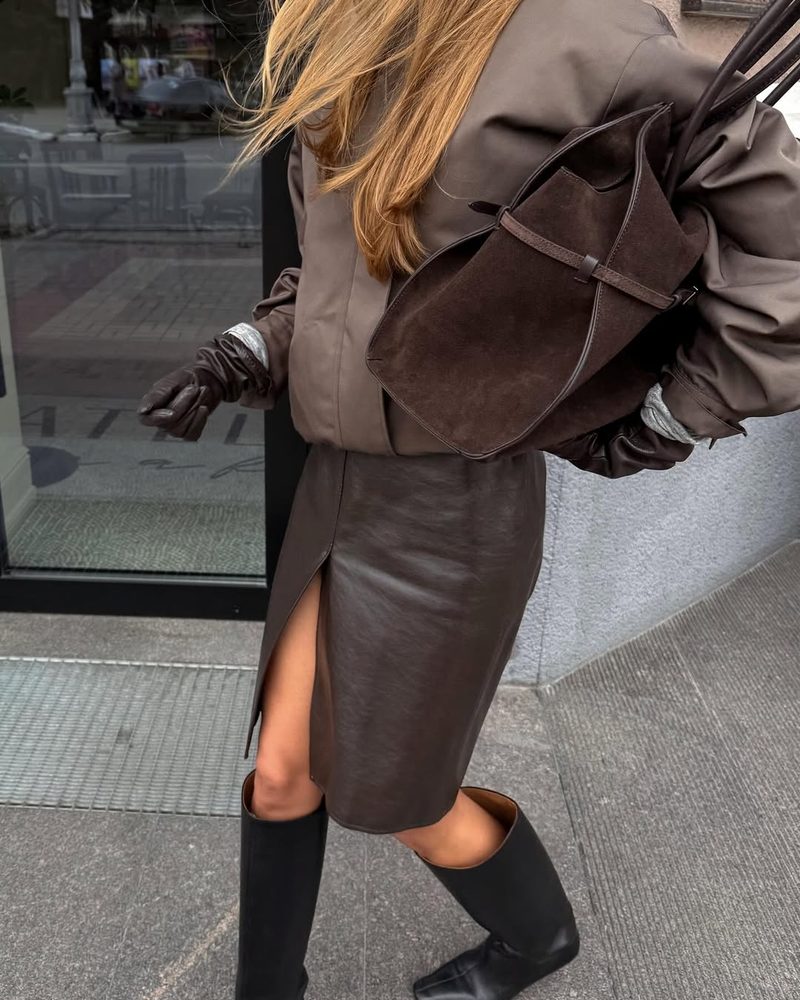 Chic urban look featuring a model in a chocolate brown leather skirt and matching cropped jacket%2C complemented by knee-high black boots and a suede handbag.