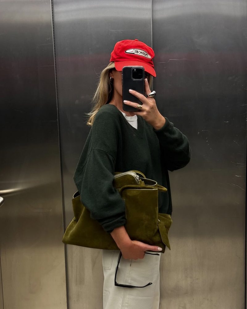 Sporty chic outfit featuring a dark green oversized sweater paired with off-white high-waisted trousers%2C complemented by a stylish olive suede tote bag and a red Nike cap