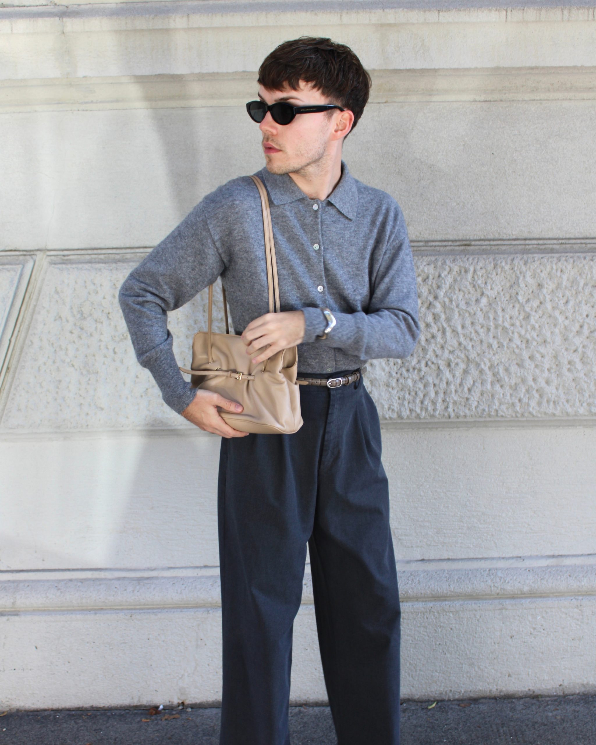 Stylish man in a grey knit polo shirt paired with tailored dark trousers%2C holding a beige leather bag and wearing sunglasses