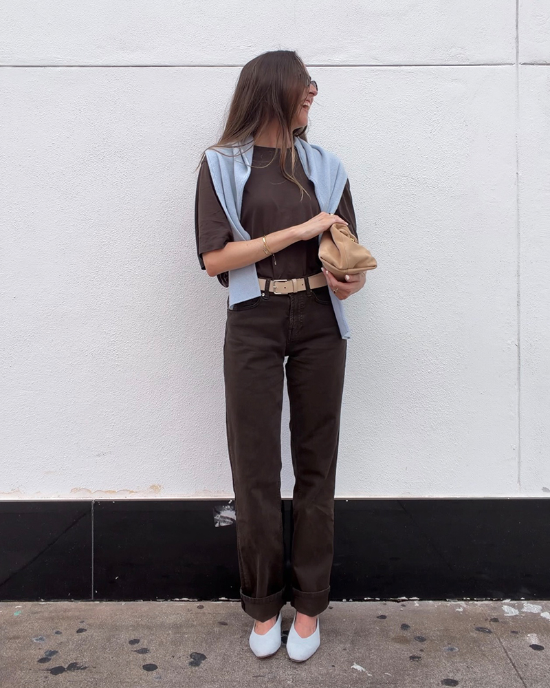 A woman wearing a brown T-shirt and dark brown jeans with a light blue sweater draped over her shoulders poses against a white wall%2C accessorized with beige flats and a tan clutch for a casual-chic street style look.