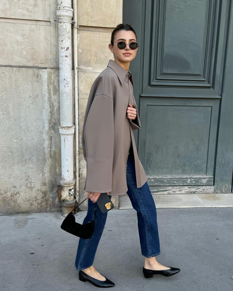 Street style look featuring an oversized taupe shirt with blue jeans%2C black flats%2C and round sunglasses%2C accessorized with a small black handbag.