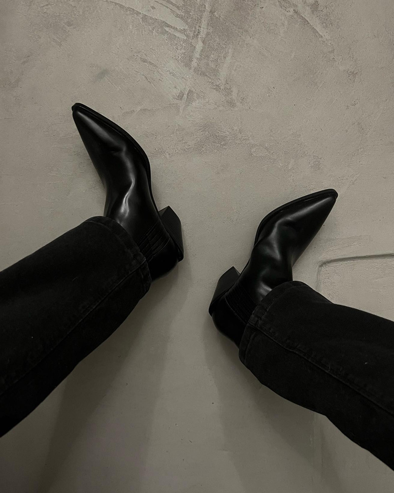Black pointed-toe leather ankle boots with low block heels styled with black jeans against a textured gray floor