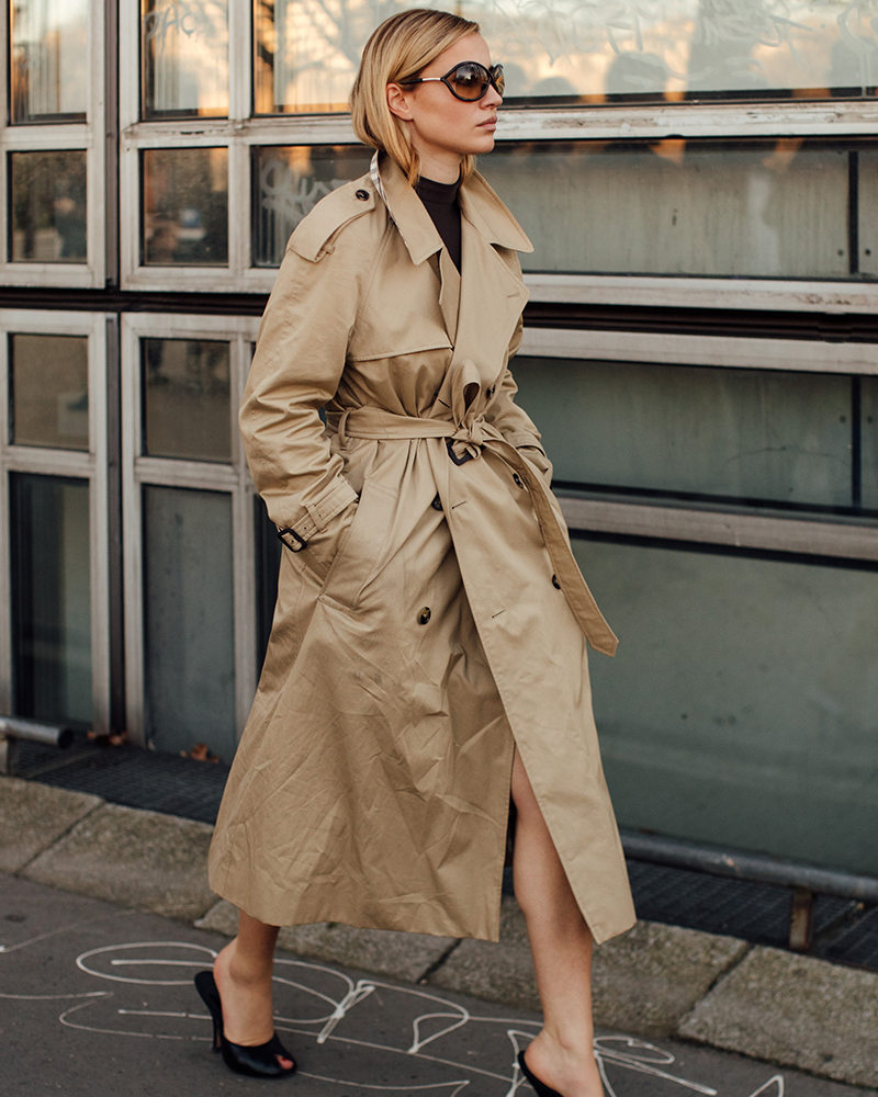 Model in a belted beige trench coat%2C black heels and oversized sunglasses walking along a city sidewalk
