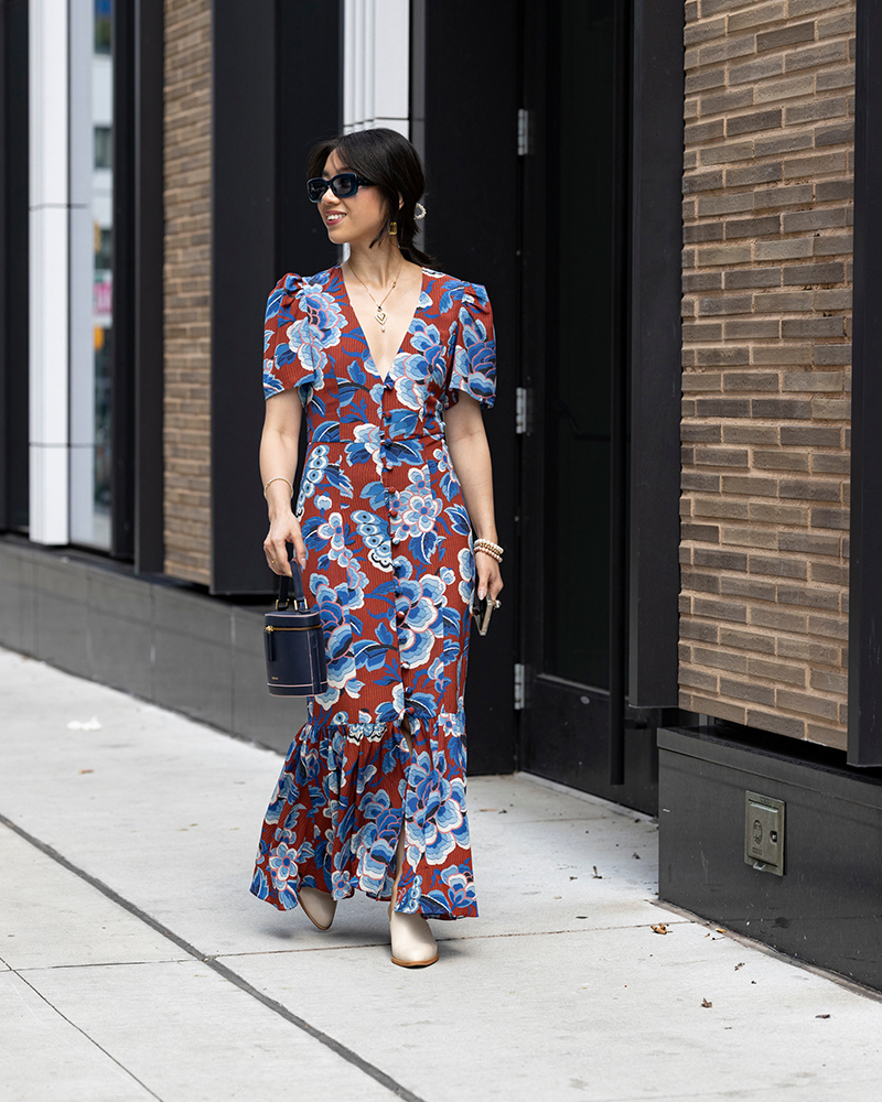Model in a red and blue floral maxi dress with sunglasses and a structured handbag on a city sidewalk
