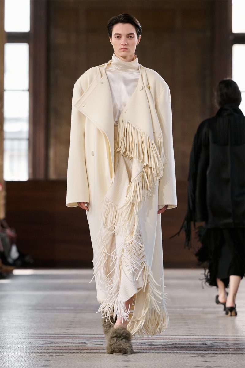 Runway model in ivory fringe coat layered over a high-neck satin dress with fuzzy slippers, walking on an indoor fashion runway backdrop