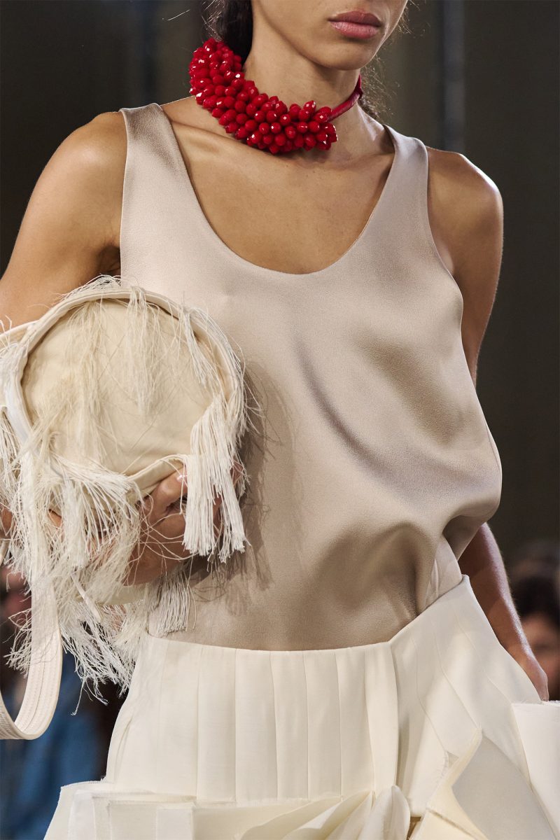 Runway look featuring ivory satin sleeveless top and pleated skirt with red beaded statement necklace and fringe clutch on fashion runway backdrop