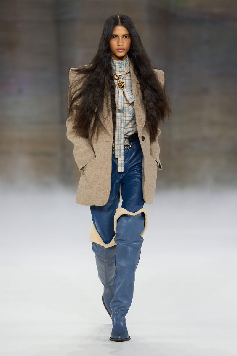 Runway look with beige wool blazer over plaid tie-neck blouse and blue leather pants, styled with knee-high boots on fashion runway backdrop
