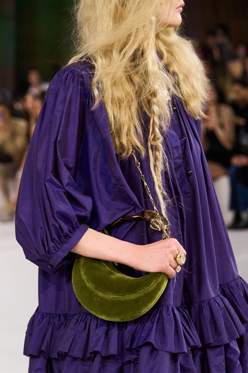 Runway look featuring a voluminous purple blouse styled with olive velvet shoulder bag and gold jewelry on fashion show runway backdrop