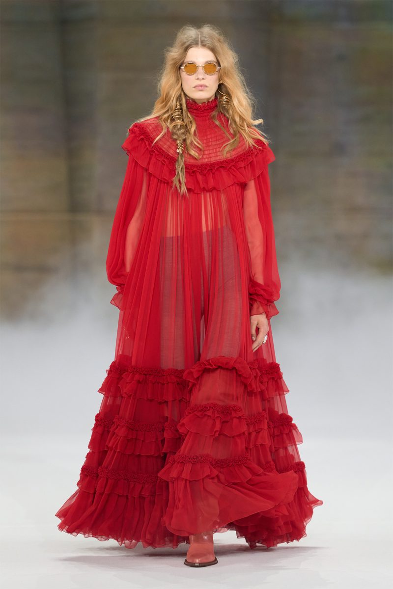 Runway look featuring sheer red ruffled chiffon gown with high neckline and round sunglasses, styled with gold earrings on fashion runway backdrop