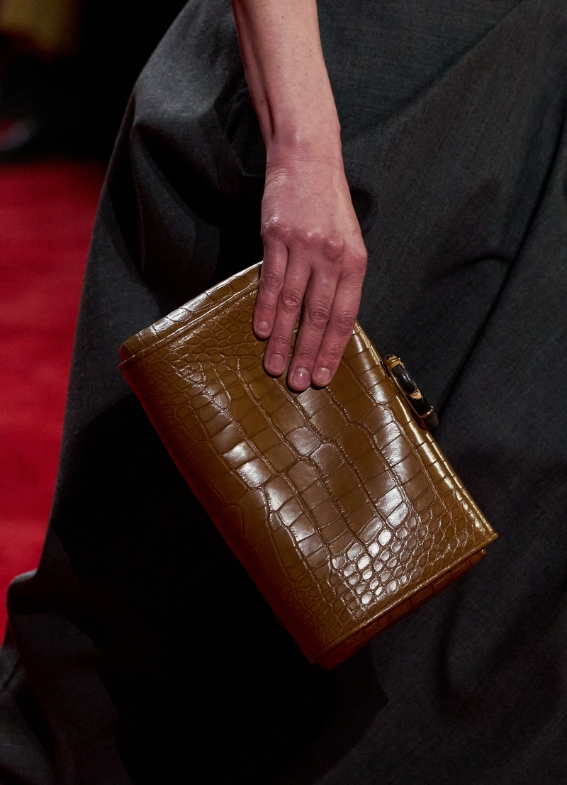 Brown croc-embossed leather clutch with glossy finish, carried against a voluminous charcoal runway dress