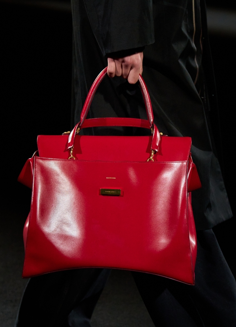 Balenciaga oversized red leather tote bag with top handles and gold-tone logo, carried with an all-black runway look