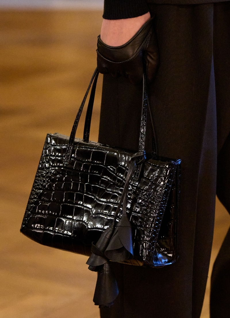 Black croc-embossed tote bag with slim handles and draped leather detail, styled with black tailoring on the runway