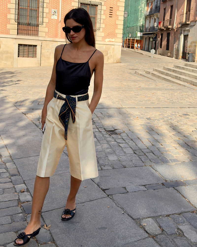 Black satin camisole styled with cream Bermuda shorts and embellished sandals on a sunlit cobblestone street