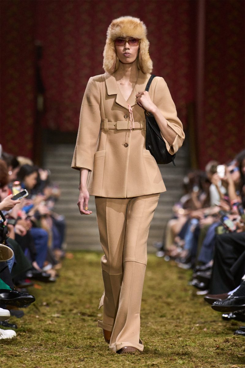 Runway model in beige belted wool suit with wide-leg trousers, fur trapper hat, sunglasses, and black shoulder bag on runway catwalk backdrop