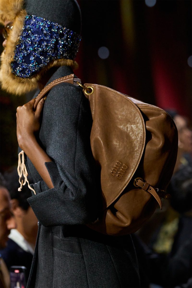 Miu Miu brown leather shoulder bag with embossed logo, styled with fur trapper hat and wool coat on runway catwalk backdrop