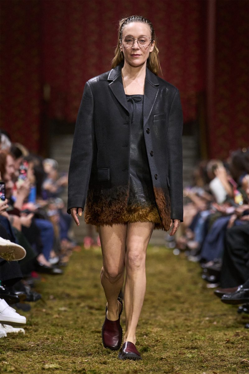 Runway model in black leather jacket and mini dress with fur trim, styled with round eyeglasses and loafers on fashion runway catwalk