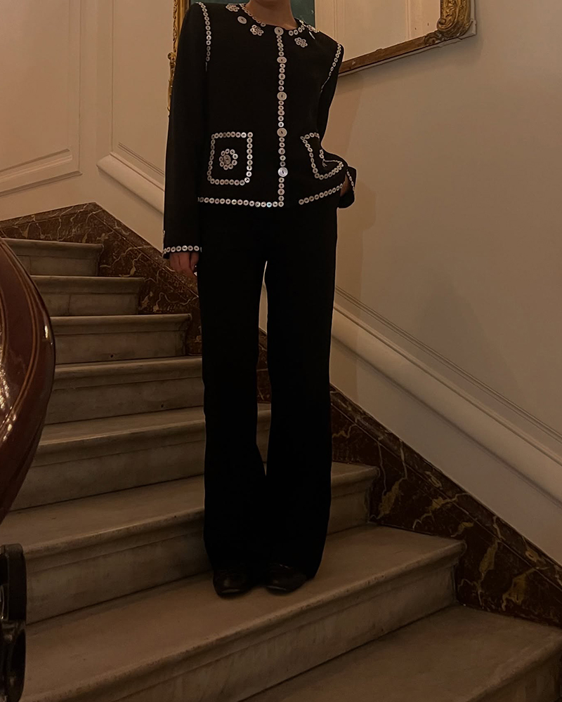 Model wears a black tailored jacket and wide-leg trousers with white trim details%2C styled on a marble staircase in a classic interior setting